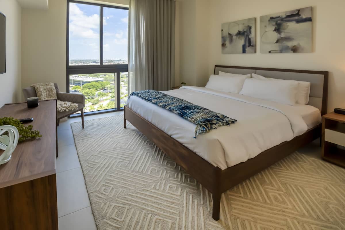 Quarto de hotel com cama grande, escrivaninha, cadeira e vista da cidade por uma janela ampla.