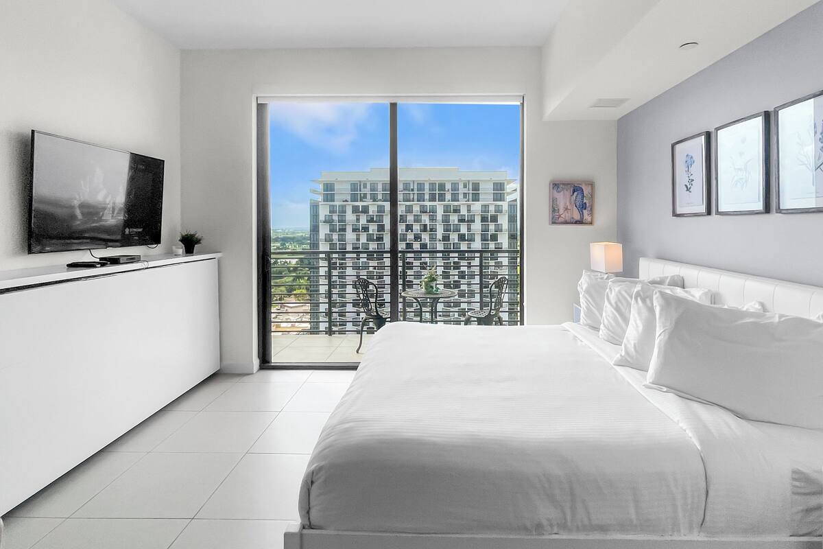Quarto de hotel moderno com uma cama grande, televisão de tela plana, varanda com vista para a cidade e uma pequena mesa com cadeiras.
