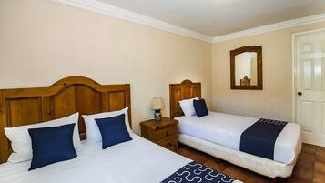 Quarto de hotel com duas camas, cabeceiras de madeira, roupa de cama branca e uma pequena mesa de cabeceira com abajur.