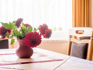Uma mesa com um vaso de flores vermelhas, uma cadeira de madeira e uma janela com cortinas.