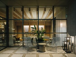 Um lobby moderno com paredes de vidro, área de estar com cadeiras e uma mesa, e uma grande planta em vaso.