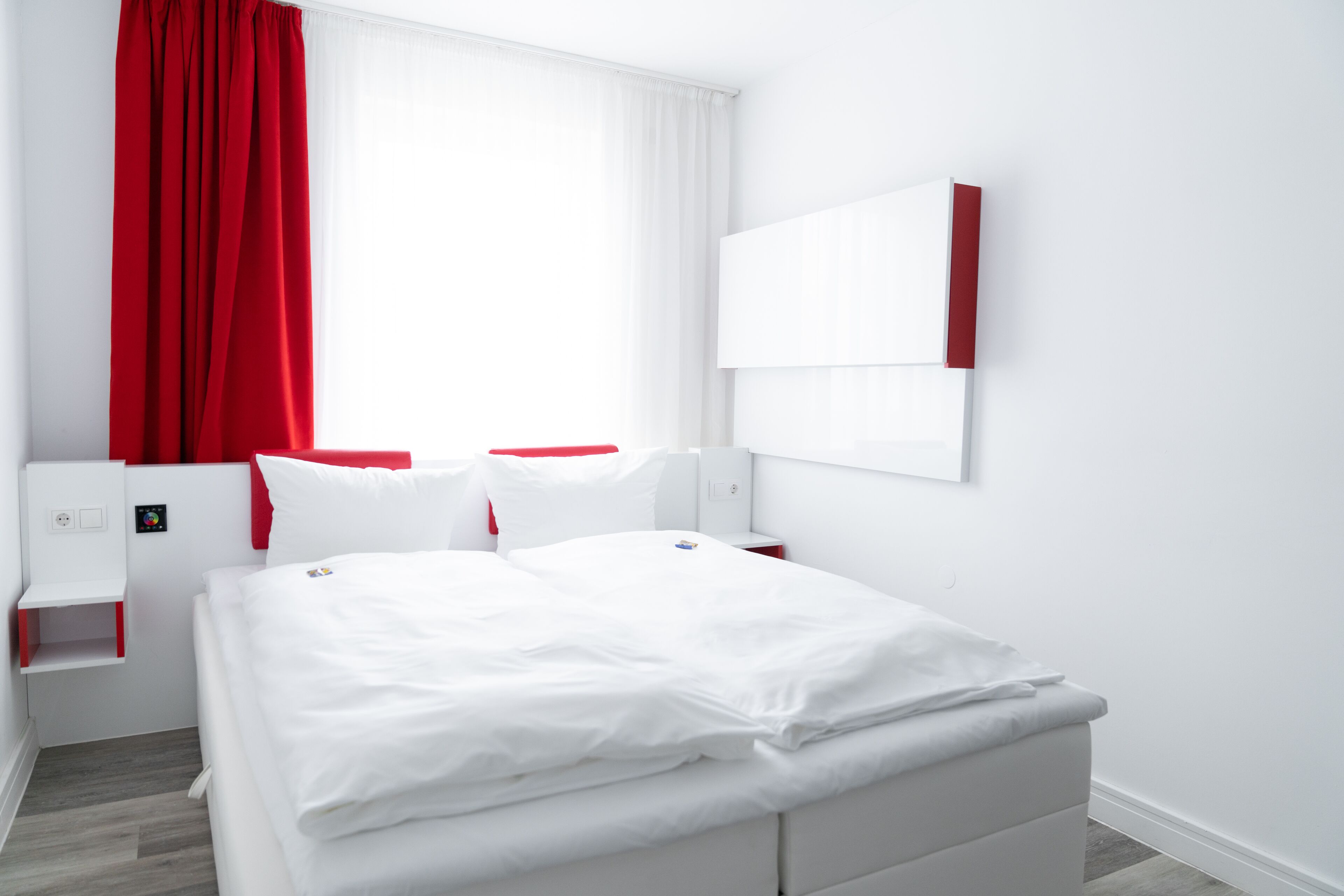Quarto de hotel moderno com uma cama bem arrumada, cortina vermelha, mesa de cabeceira branca e luminária fixa na parede.