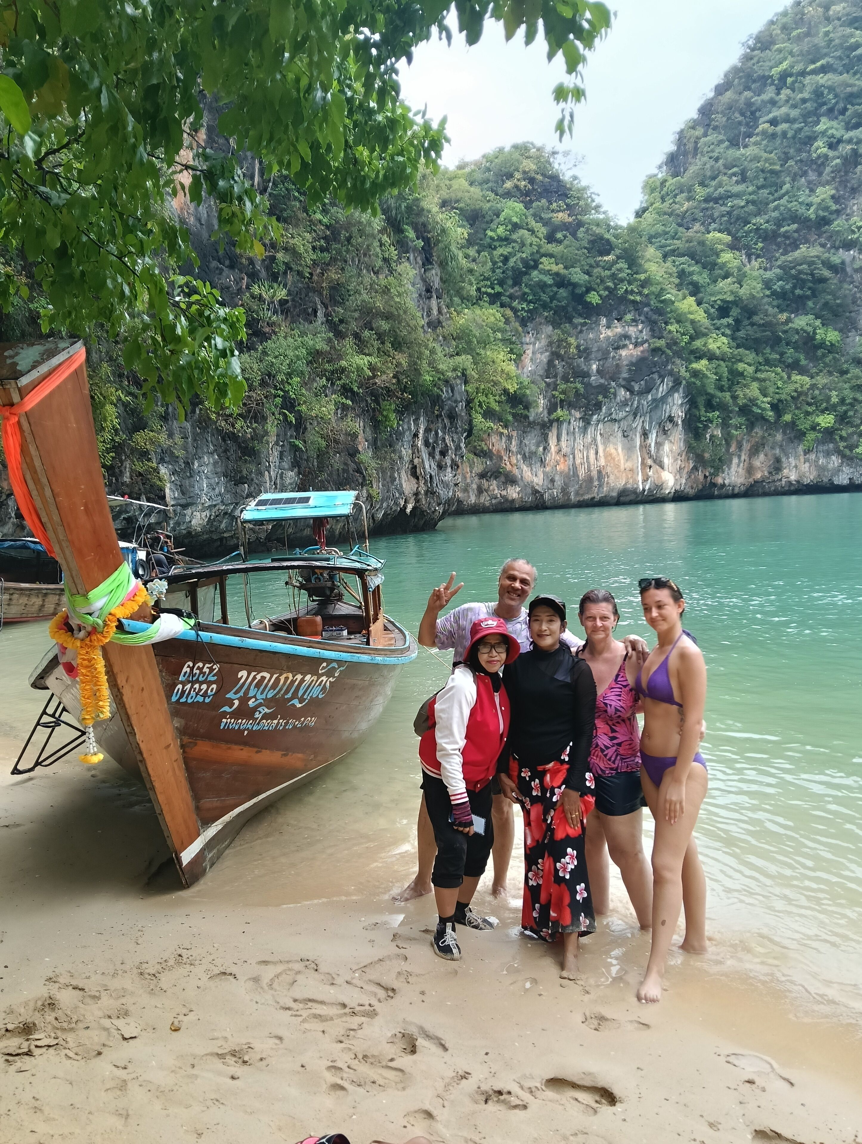 Um grupo de pessoas está em pé numa praia arenosa perto de um barco, com um penhasco rochoso ao fundo.