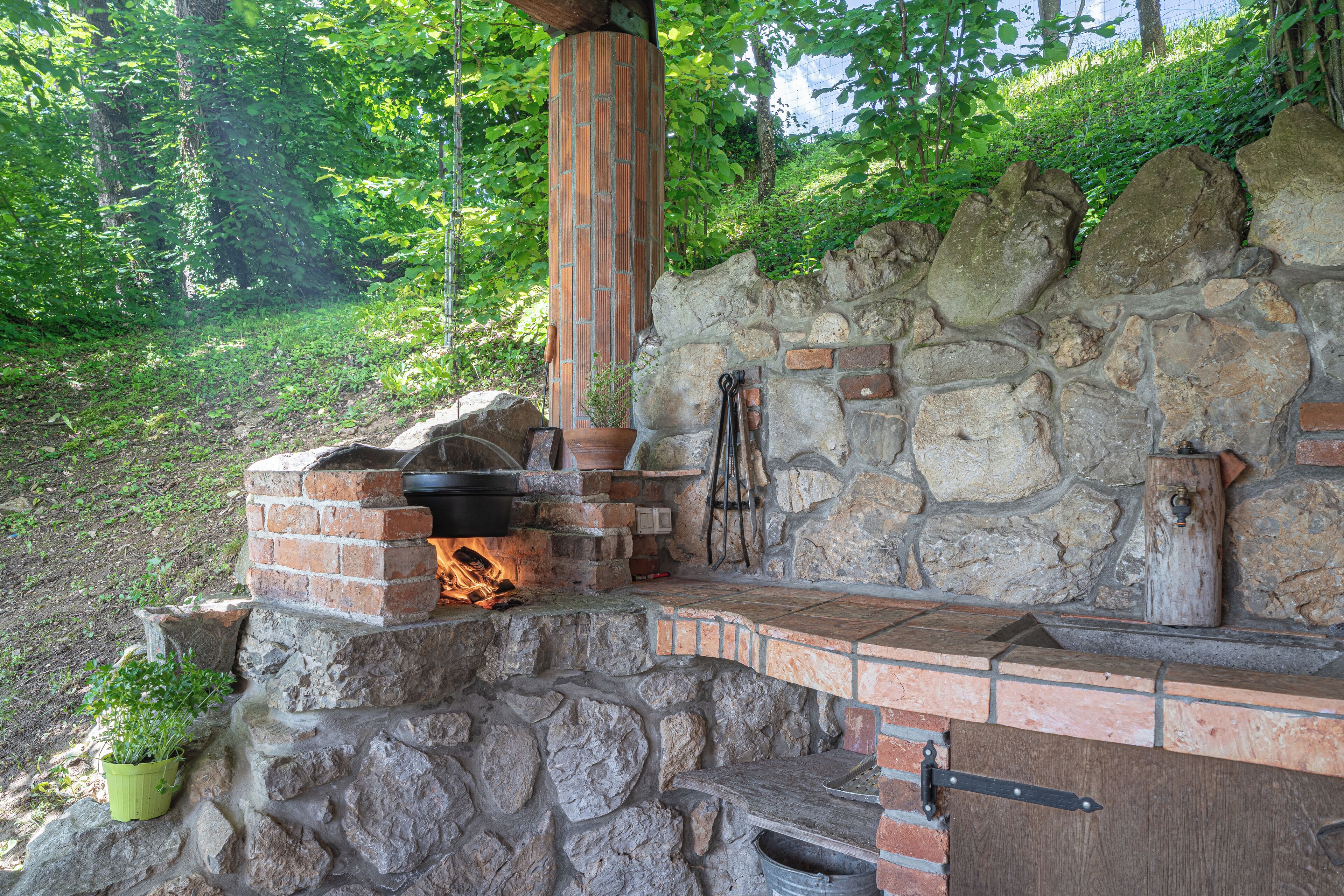 Lareira de pedra ao ar livre com forno de tijolos, cercada por um muro de pedra e vegetação.