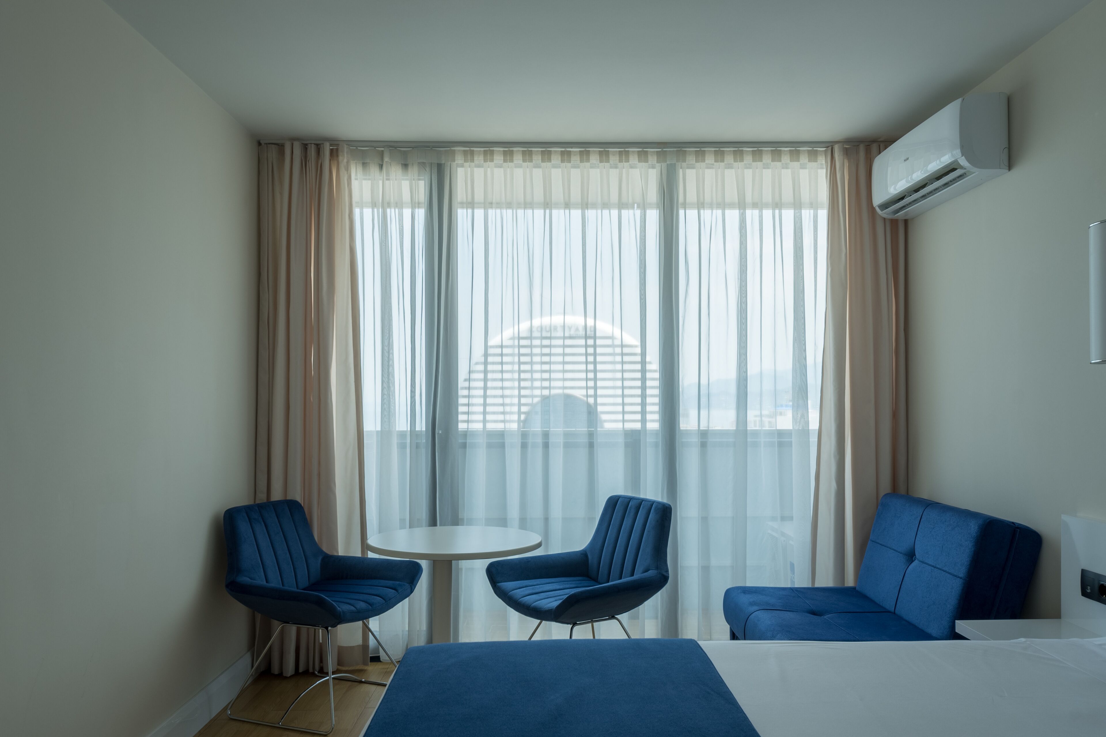 Quarto de hotel com uma cama, duas poltronas azuis, uma mesa redonda pequena e uma janela grande com cortinas leves.