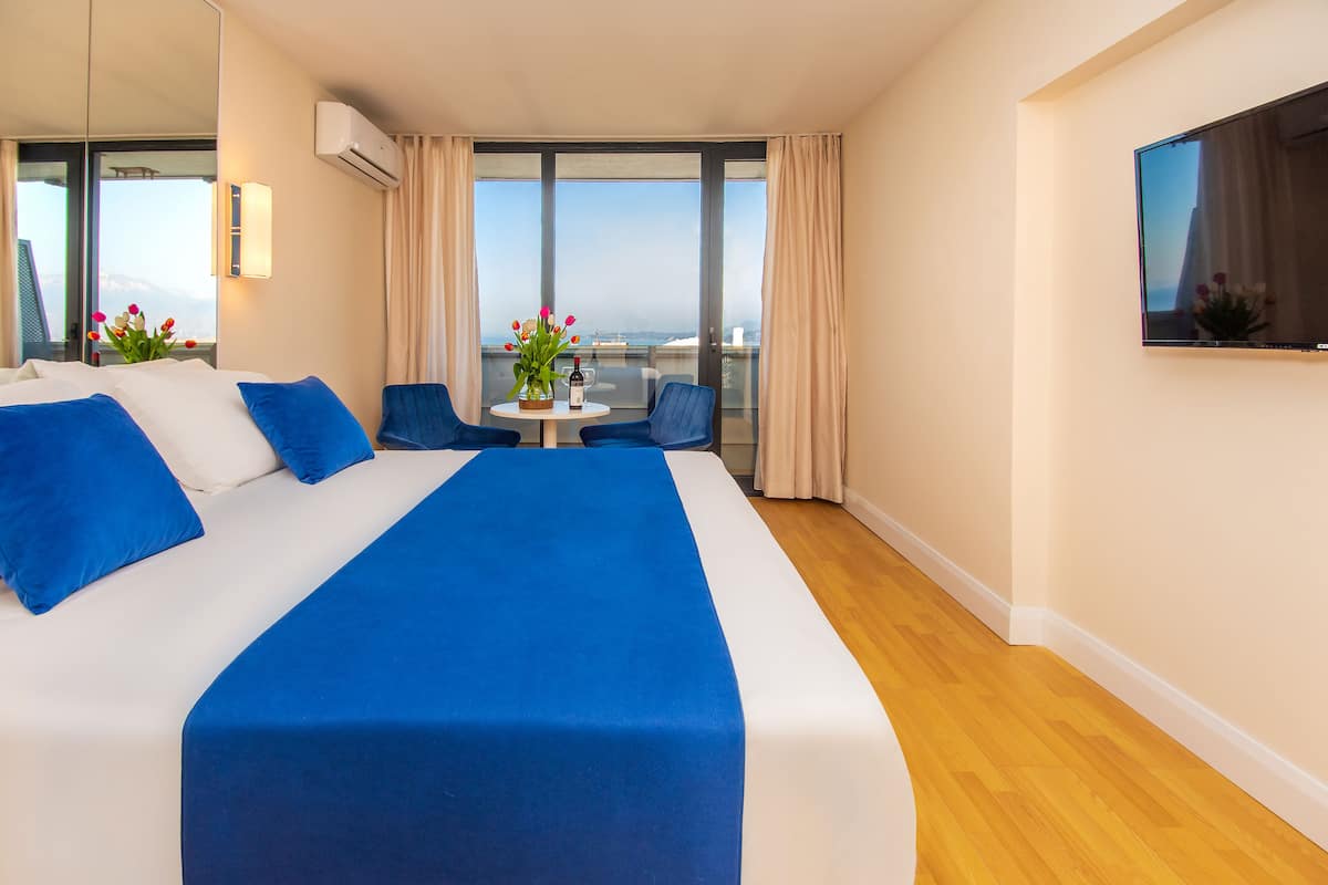 Quarto de hotel com uma cama grande, uma mesa pequena com cadeiras, uma televisão na parede e uma varanda com vista.
