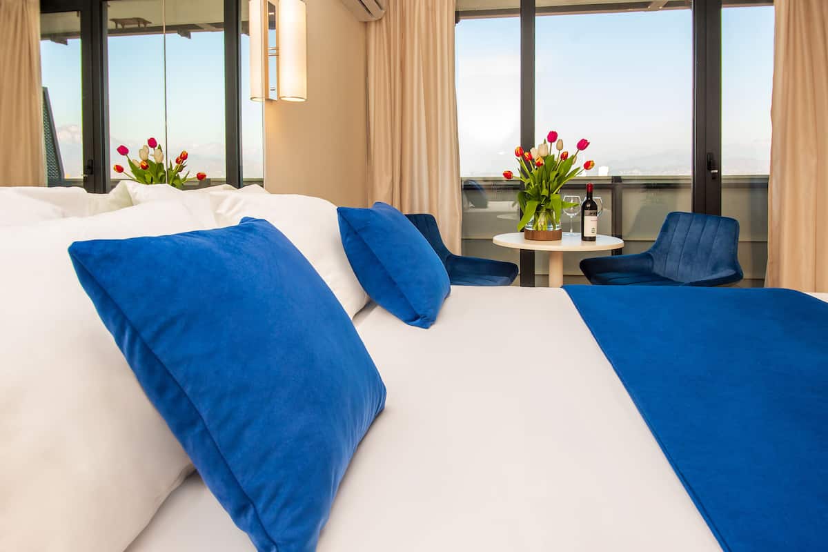 Quarto de hotel com uma cama grande, almofadas azuis, uma pequena mesa redonda com um vaso de flores e vista da cidade através de amplas janelas.