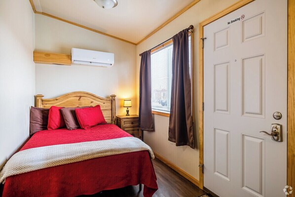 Quarto com cama de madeira, roupa de cama vermelha, mesa de cabeceira com abajur, janela com cortinas e porta com placa de 'Proibido Fumar'.