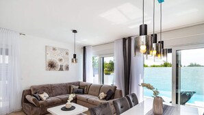 Uma sala de estar moderna com um sofá seccional, uma mesa de centro e uma área de jantar com mesa de vidro. Há uma janela grande com vista para o mar.