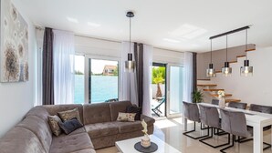Sala de estar moderna com sofá, mesa de centro e área de jantar com cadeiras. Há uma janela grande com cortinas, uma luminária no teto e vista para uma piscina e uma casa lá fora.