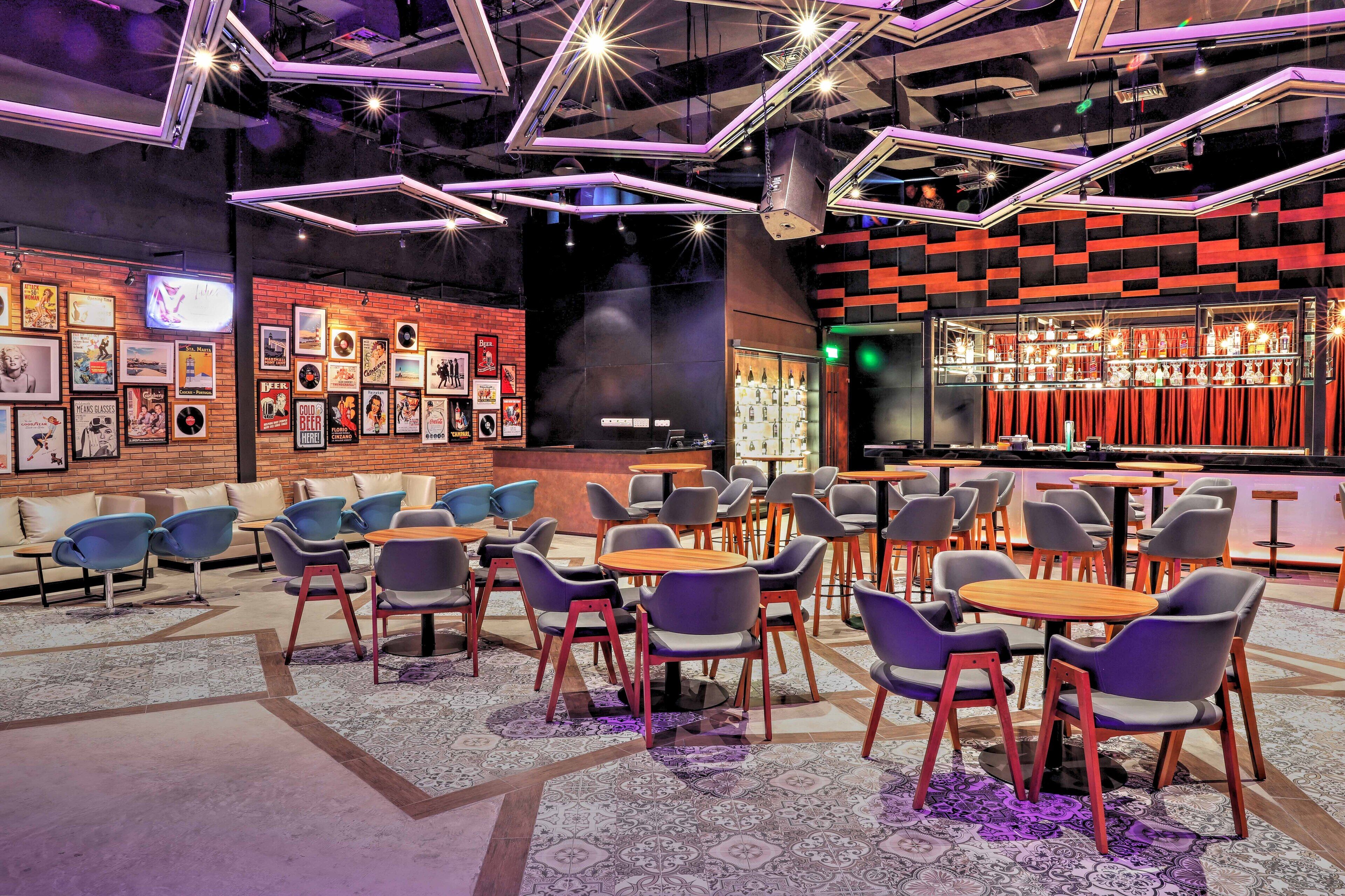 Um espaço de bar moderno com parede de tijolos, luzes de neon e diversas opções de assento, incluindo mesas e cadeiras.
