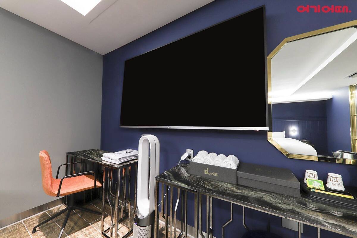 Quarto de hotel com televisão de tela plana grande fixada na parede, toucador moderno, uma cadeira e um espelho.
