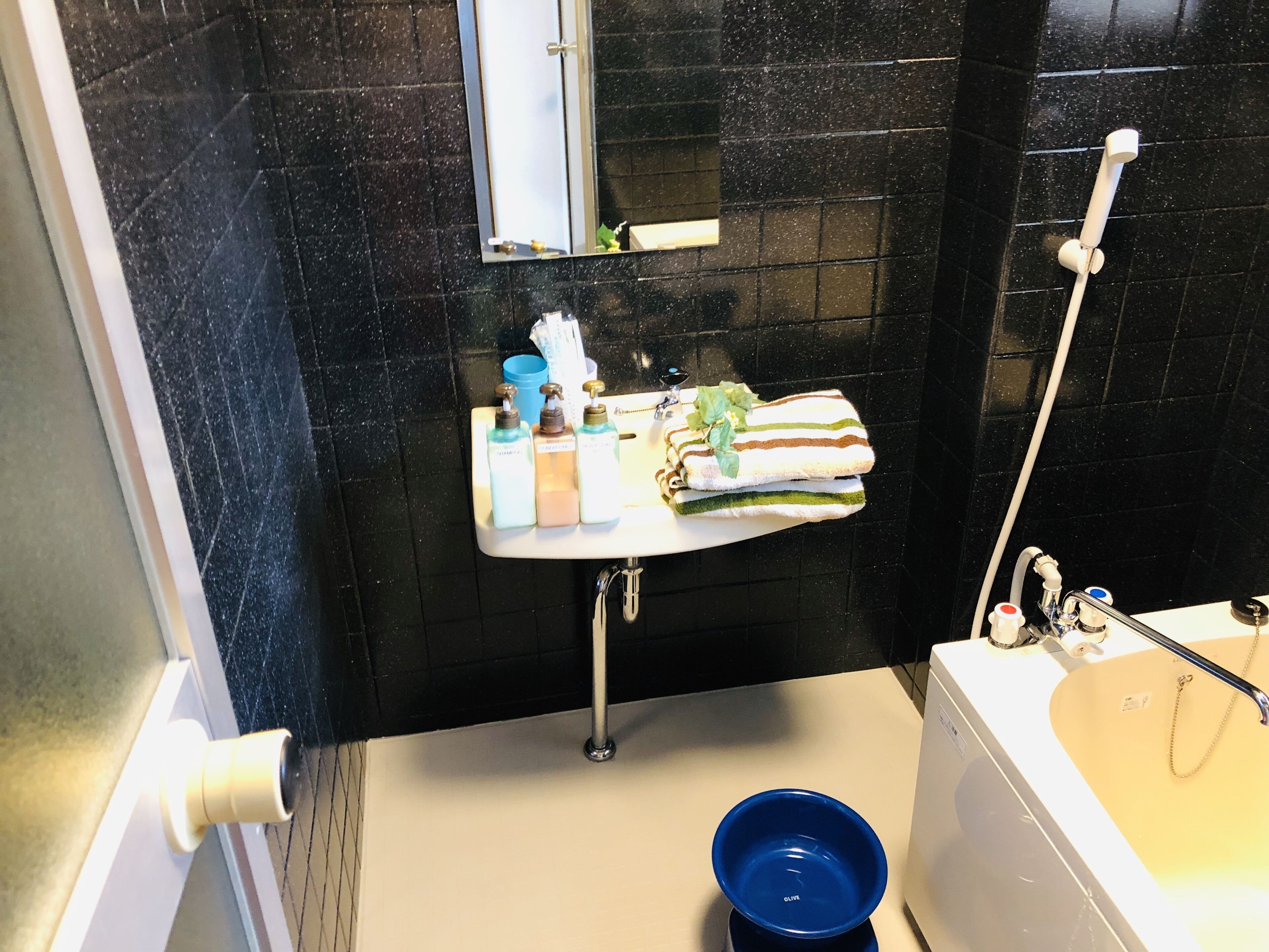 Um banheiro com parede revestida de azulejos pretos, pia branca com artigos de higiene, um recipiente azul e uma banheira.