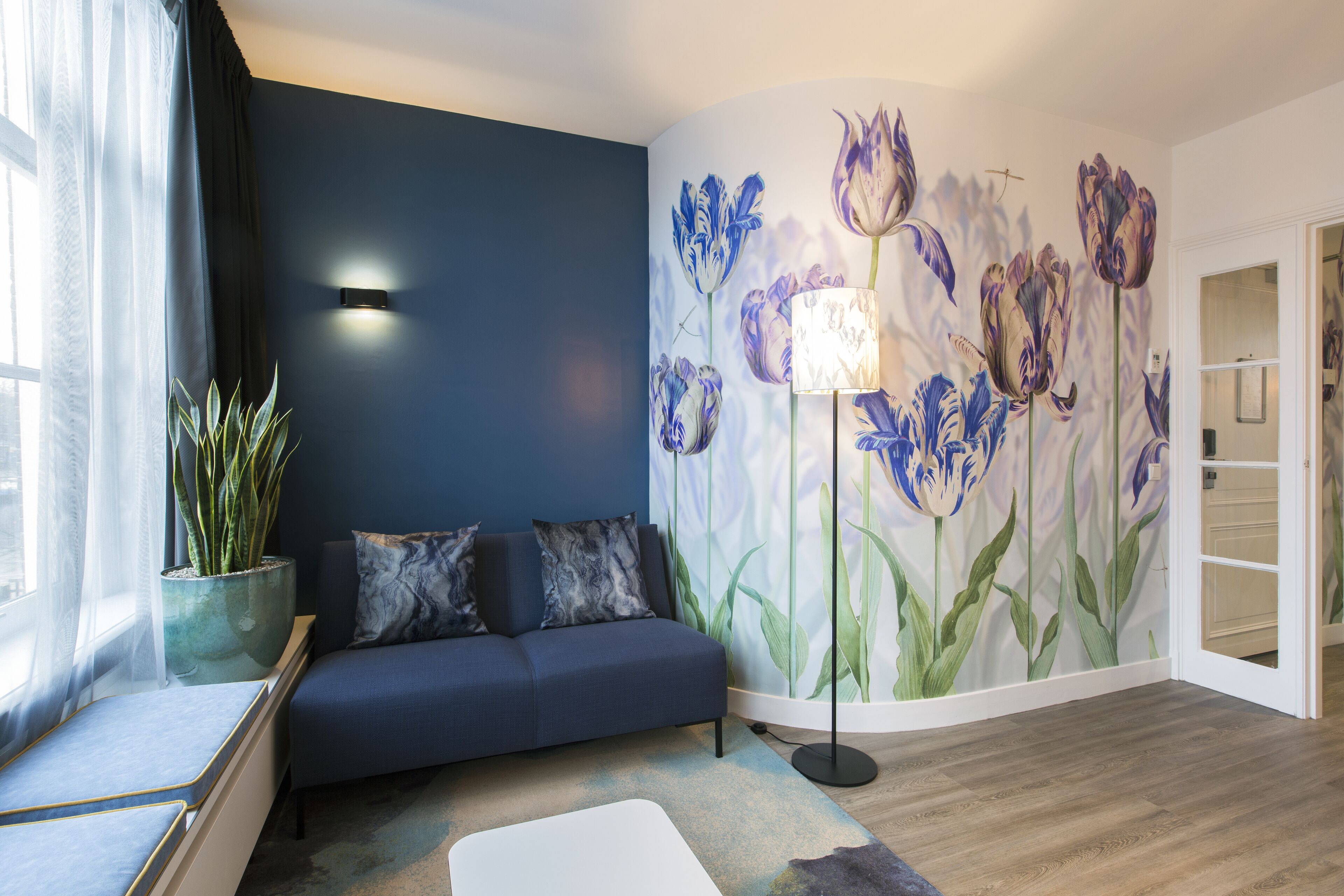 Uma sala de estar moderna com um sofá azul escuro, uma luminária de chão, uma janela grande com cortinas finas e um mural de flores azuis na parede.