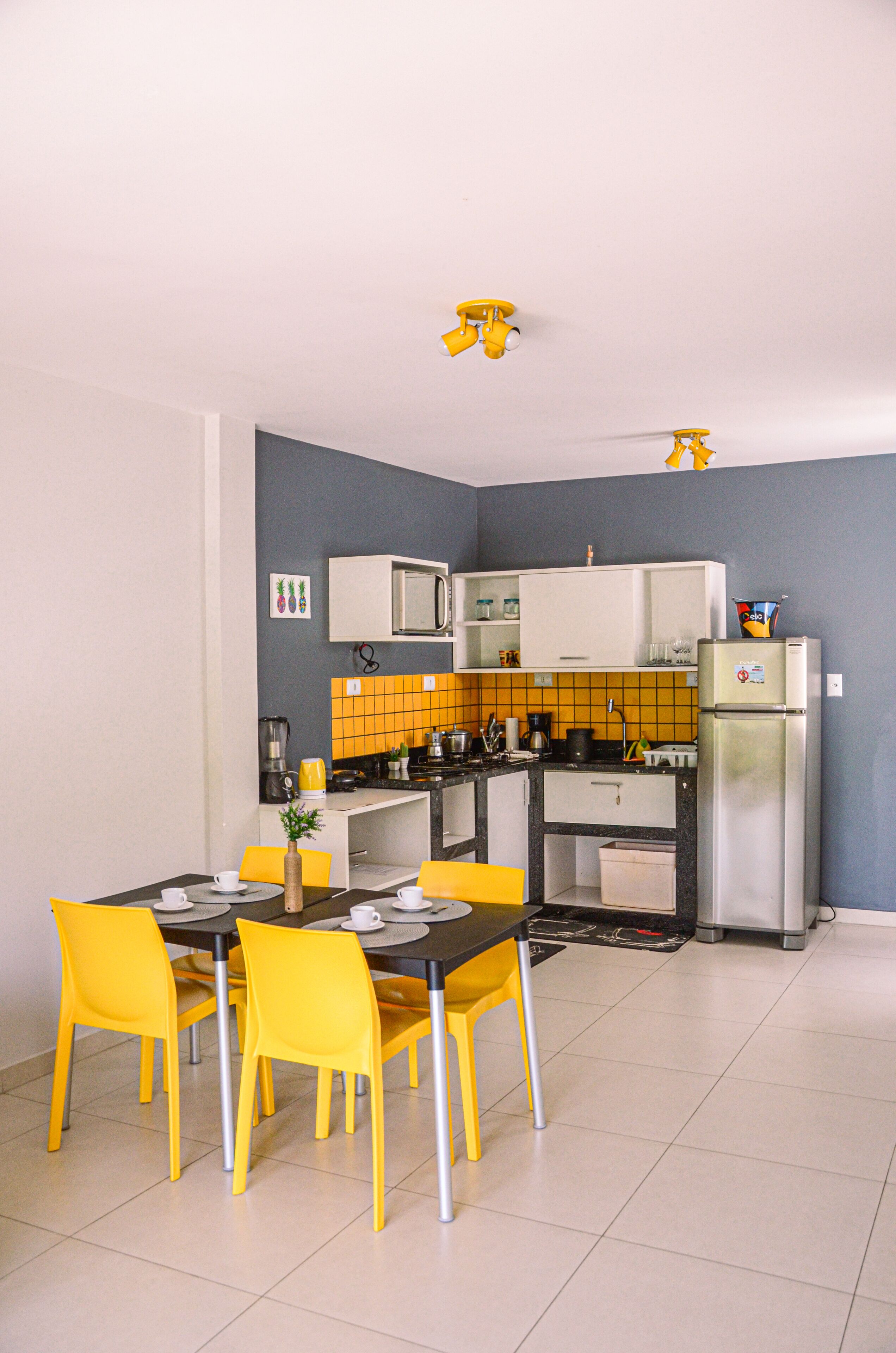 Cozinha compacta com micro-ondas, fogão e frigorífico. Há uma mesa de jantar com cadeiras amarelas e uma pequena planta.