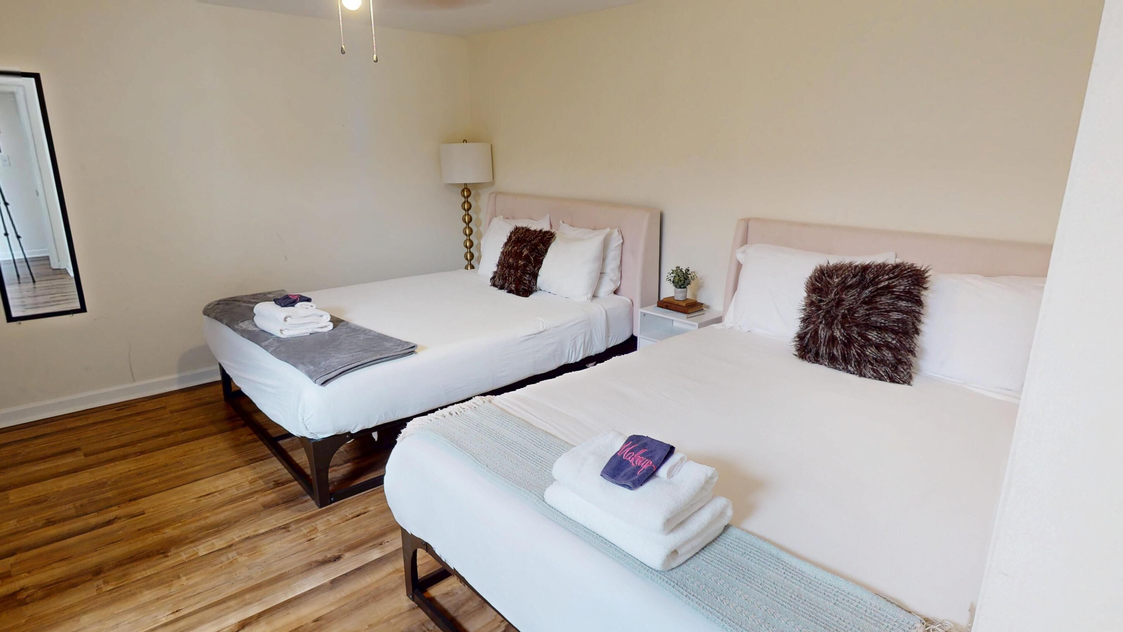 Quarto de hotel com duas camas, uma pequena mesa de cabeceira com abajur e piso de madeira.