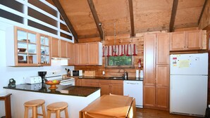 Uma cozinha com armários de madeira, uma geladeira branca, micro-ondas, fogão e uma janela com cortinas. Há uma mesa de madeira com dois banquinhos.
