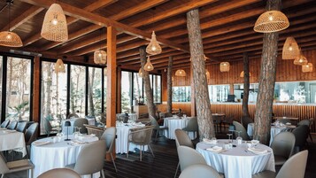 Um restaurante com mesas redondas, vigas de madeira e grandes janelas.