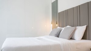 Cama bem arrumada com edredom branco e travesseiros, cabeceira cinza e luminárias fixadas na parede.