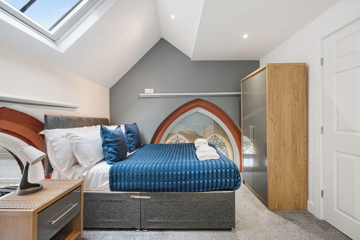 Quarto com claraboia, cama com colcha azul, guarda-roupa de madeira e porta branca.