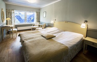 Quarto de hotel com uma cama grande, uma escrivaninha com abajur, uma cadeira e vista para montanhas pela janela.