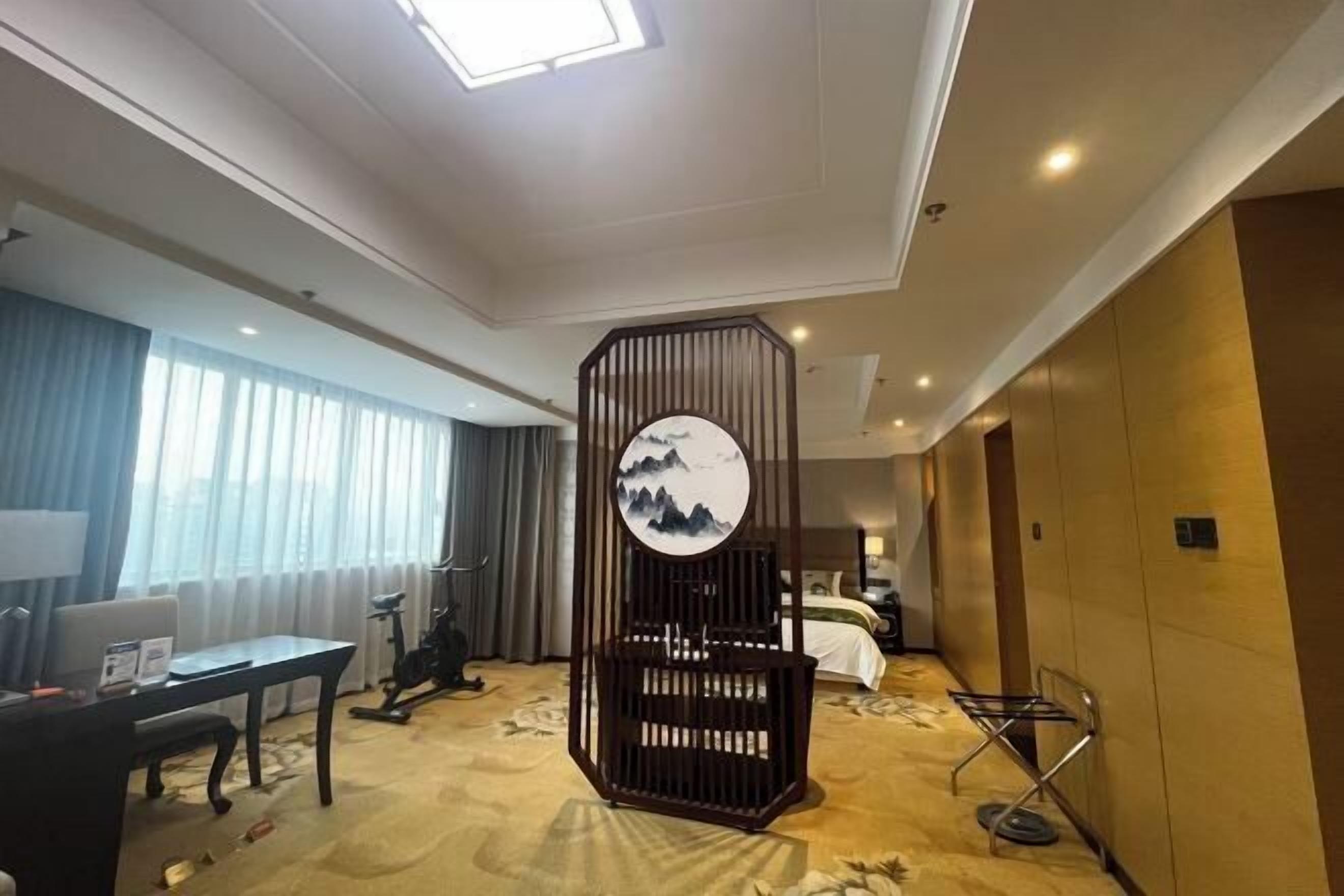 Quarto de hotel com cama, equipamentos de ginástica, escrivaninha e uma janela grande com cortinas.