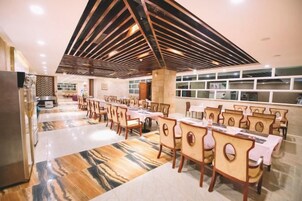 Uma sala de jantar espaçosa, com várias mesas e cadeiras, design de teto moderno e área de cozinha ao fundo.