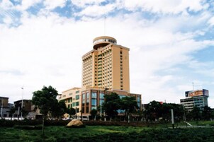 Um hotel alto e cilíndrico com um detalhe circular central, cercado por vegetação e outros edifícios.