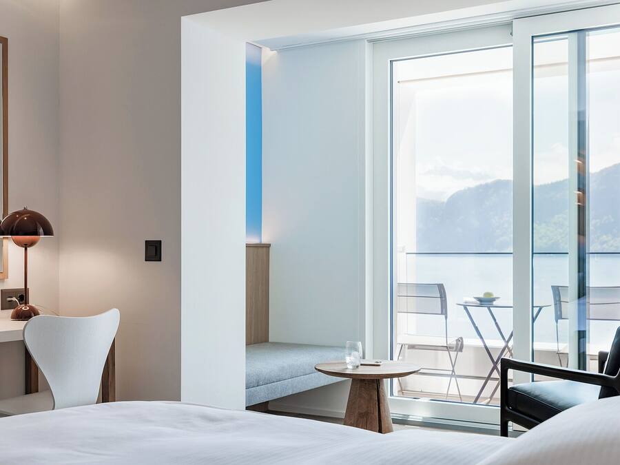 Quarto de hotel moderno com uma grande janela, área de estar com mesa e escrivaninha com luminária.