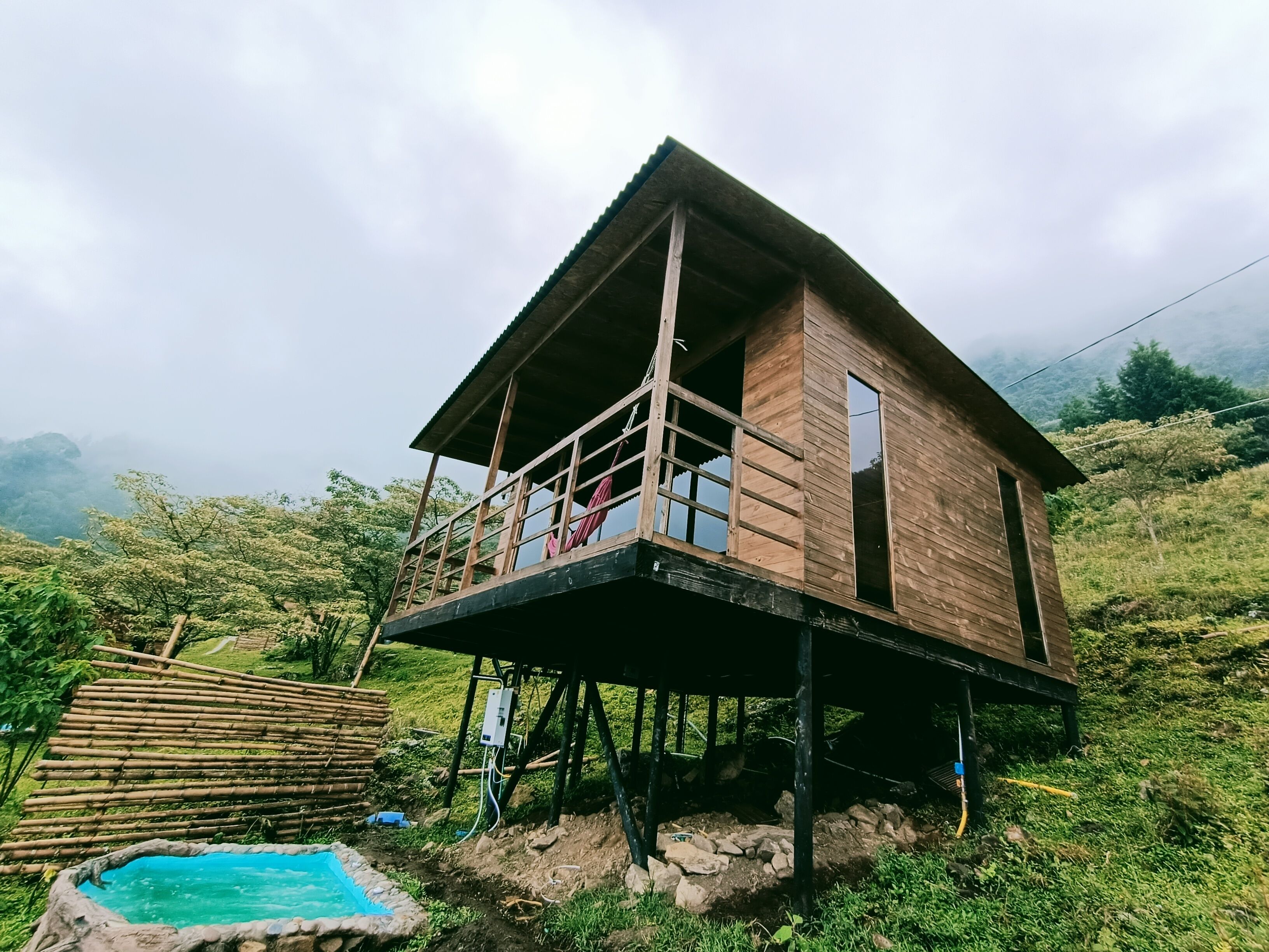 Uma casa de madeira com varanda, erguida sobre pilares, cercada por vegetação e uma piscina pequena.