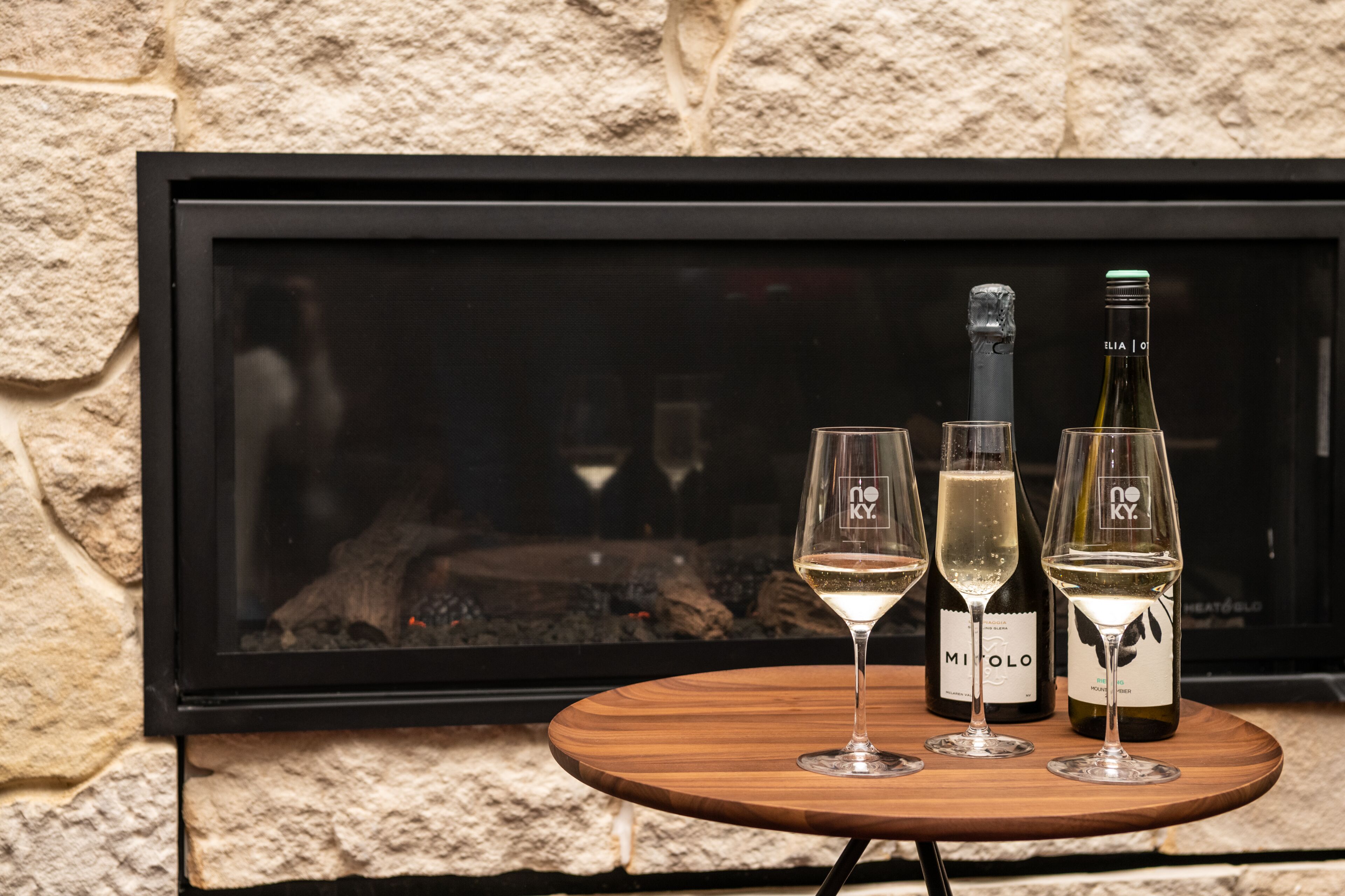 Uma mesa de madeira com duas taças e duas garrafas de vinho, em frente a uma parede de pedra e uma lareira.