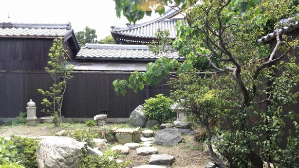 Um jardim japonês tradicional com caminhos de pedra, uma pequena árvore e um edifício de madeira com telhado de telhas.