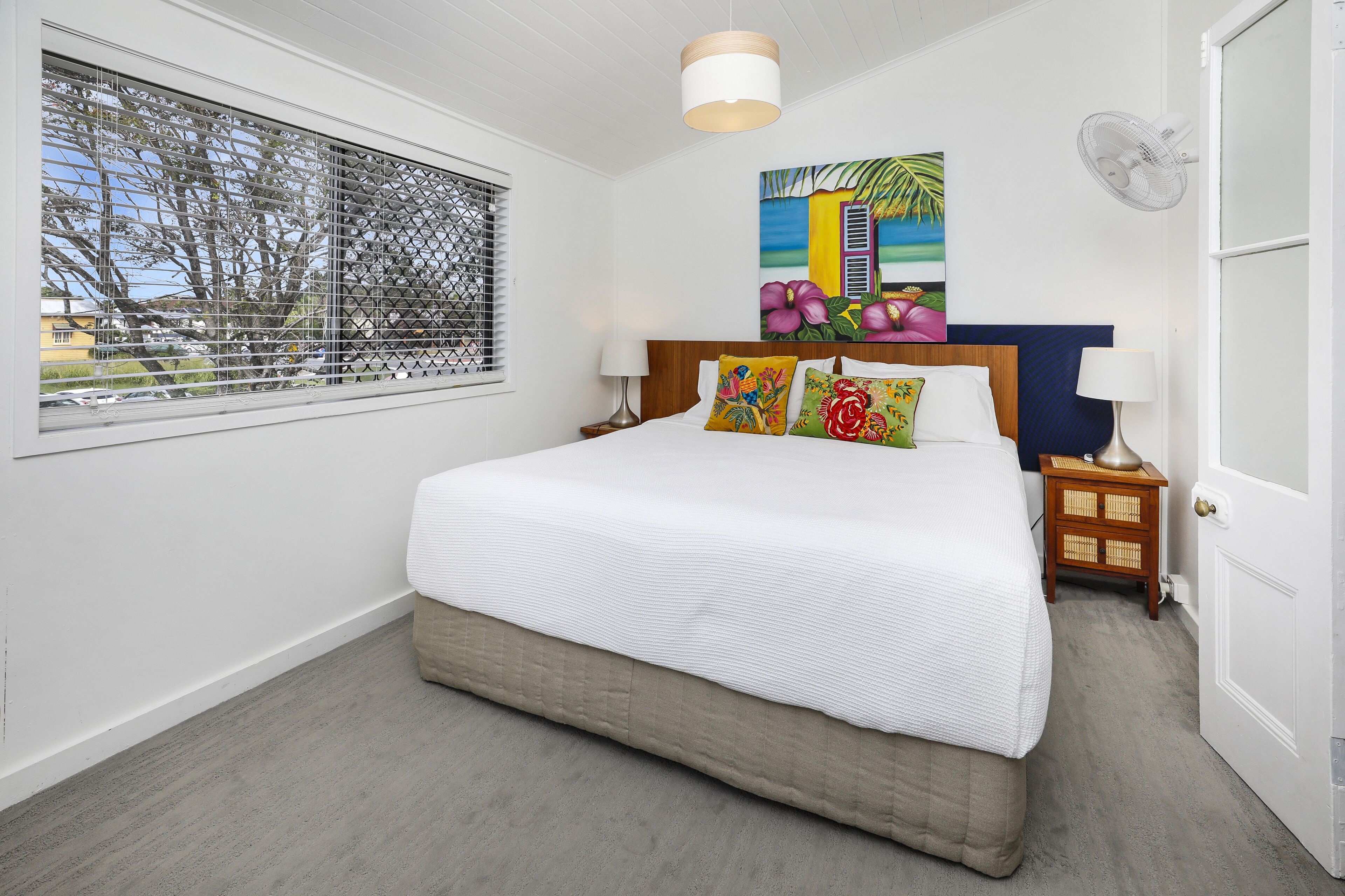 Quarto com cama grande, janela com persianas, um quadro na parede e mesa de cabeceira com luminária.