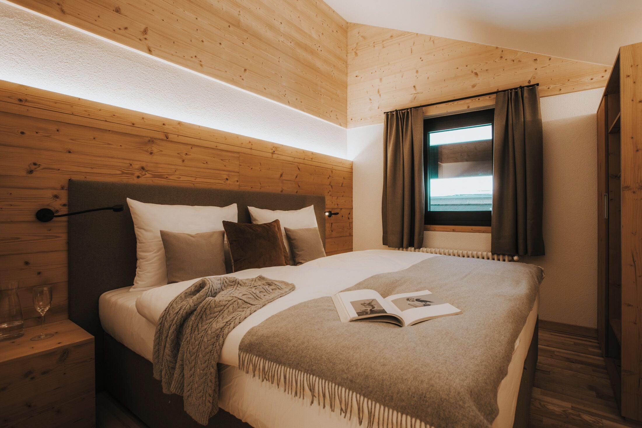 Quarto com cabeceira de madeira, cama grande com cobertor, mesa de cabeceira com um copo e janela com cortinas.