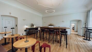 Um quarto amplo com uma longa mesa de madeira e vários banquinhos, teto branco com uma cruz e vista para outro cômodo por uma porta aberta.