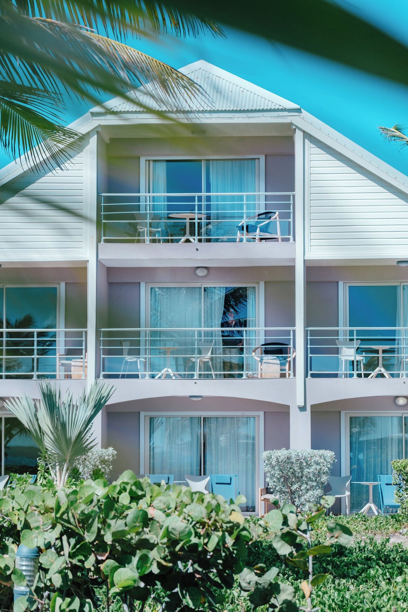 Um hotel de vários andares com varandas, janelas amplas e um jardim tropical.