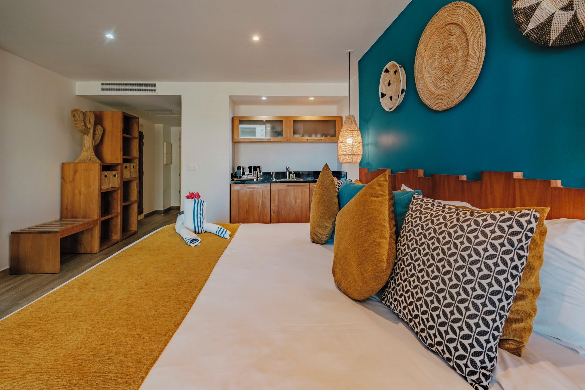 Quarto de hotel com cama, almofadas, cabeceira de madeira, área de cozinha e banheiro ao fundo.