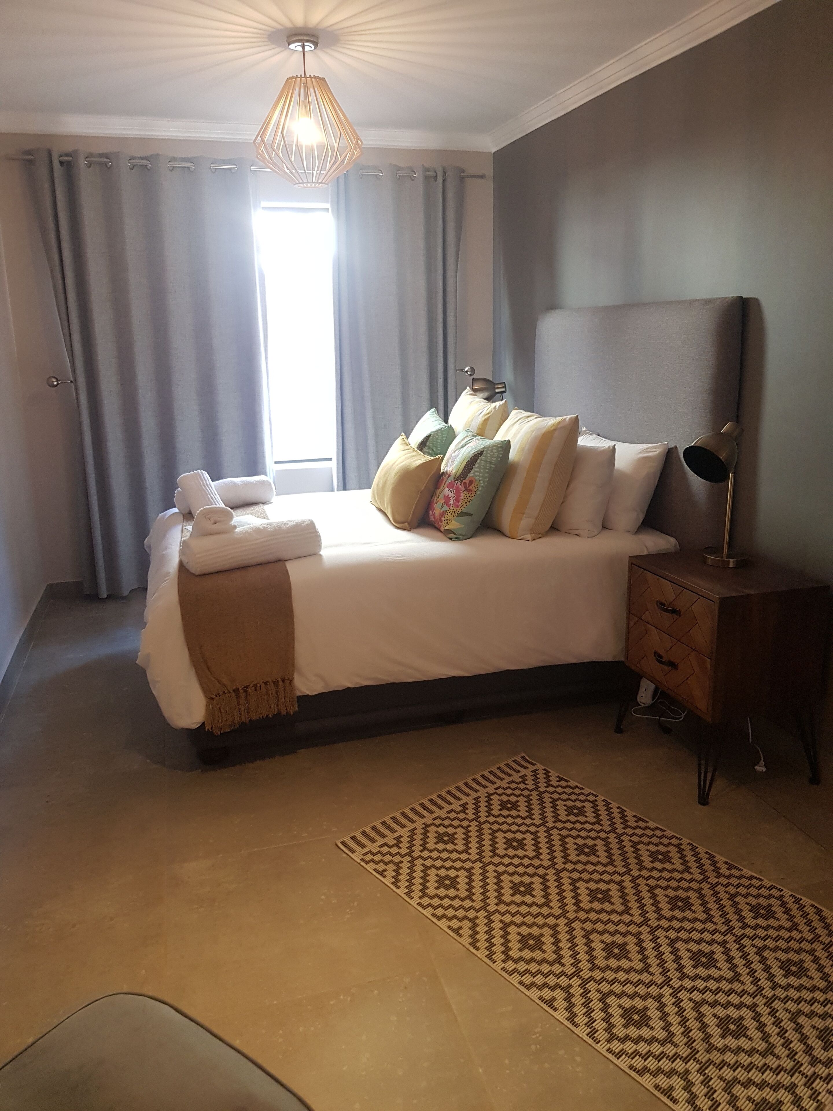 Uma cama bem arrumada com cabeceira, mesa de cabeceira com abajur e tapete estampado sobre piso de azulejos.