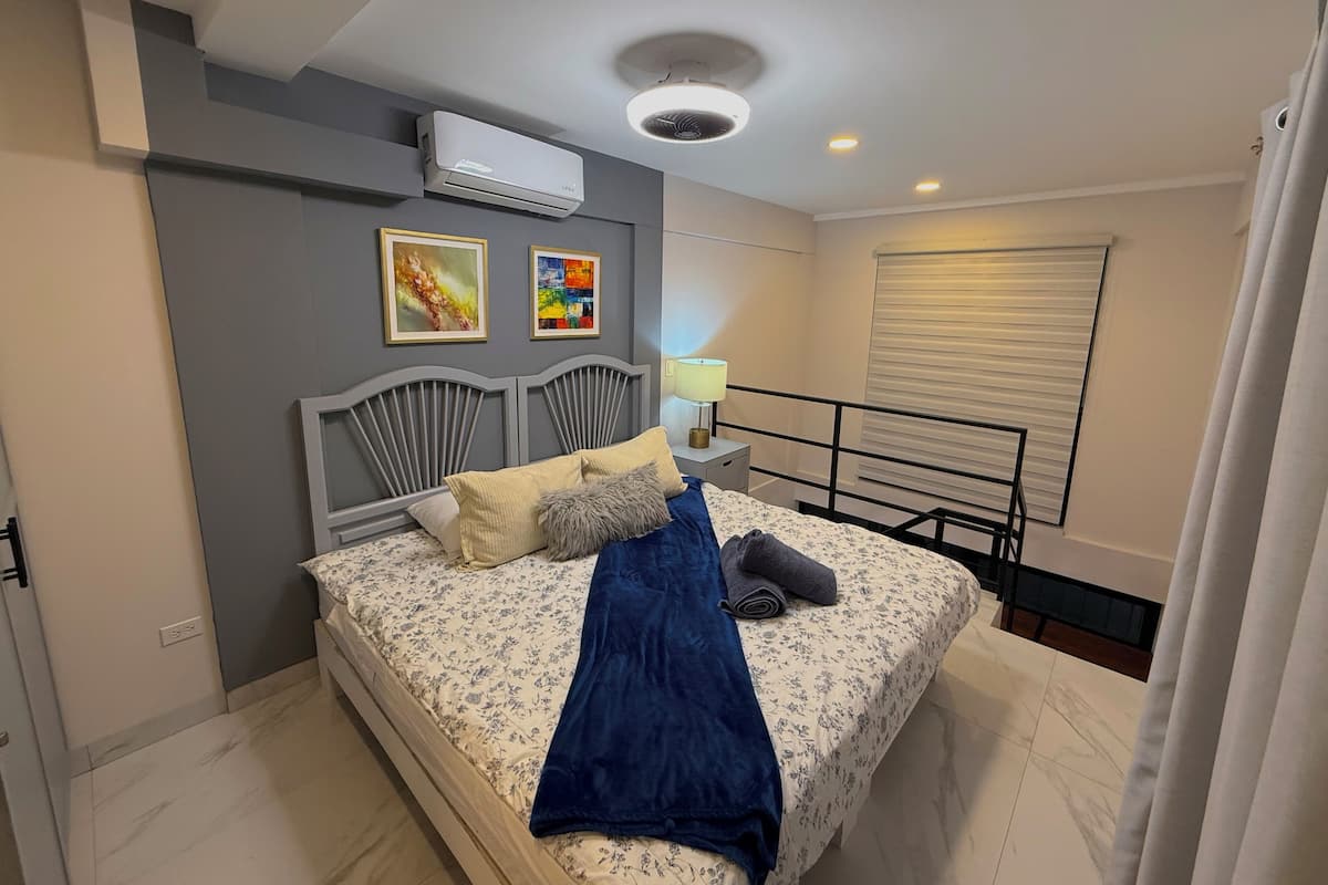 Um quarto com cama, mesa de cabeceira, luminária, ar condicionado e janela com persianas.