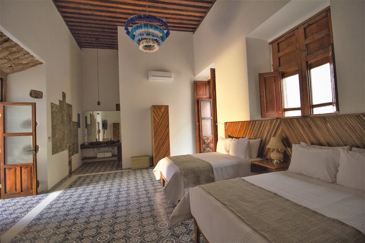 Quarto com duas camas, teto de madeira, ladrilhos no chão com padrão, um candelabro e uma janela com persianas de madeira.