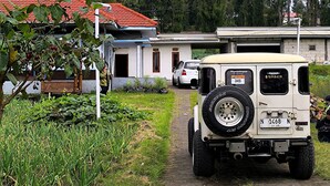 Um SUV branco estacionado em frente a uma casa com telhado vermelho, cercado por vegetação e árvores.