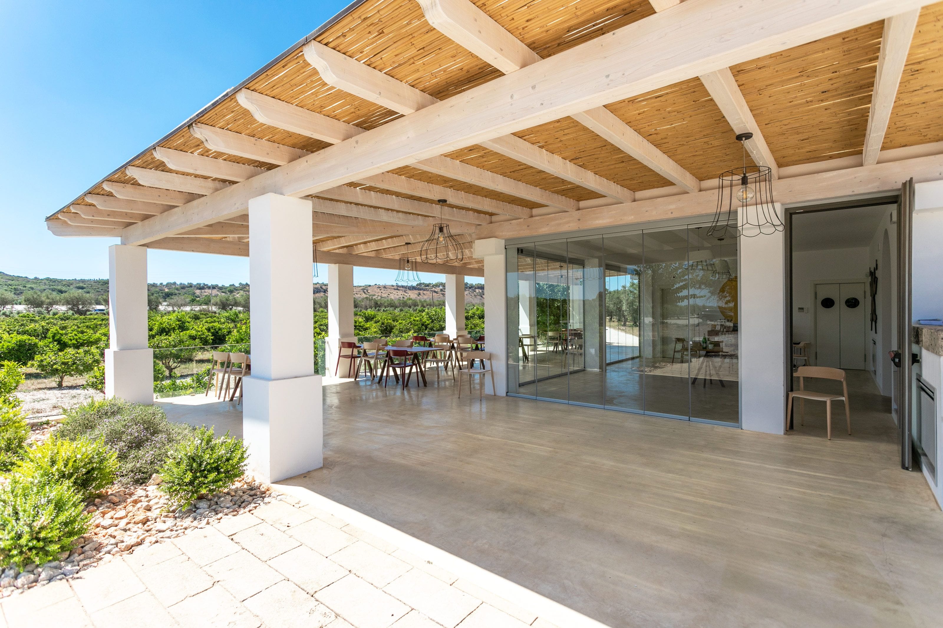 Um pátio externo com pérgola de madeira, portas de vidro que dão para uma cozinha e vista para um vinhedo.