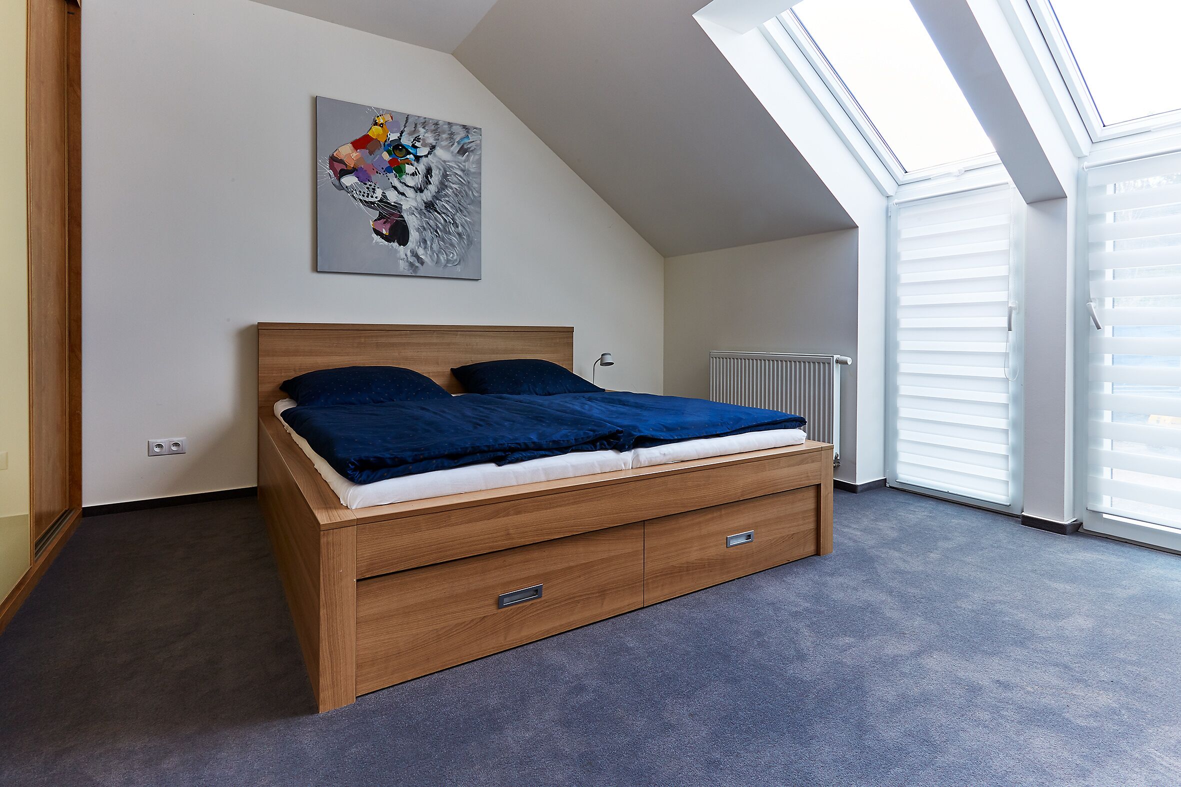 Quarto com teto inclinado, cama com estrutura de madeira, radiador e claraboia.