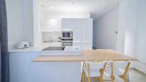 Cozinha moderna com armários brancos, micro-ondas e torradeira. Há uma mesa de jantar de madeira com cadeiras brancas.