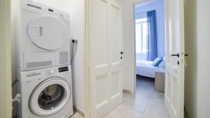 Uma área de lavanderia com máquina de lavar e secadora, um quarto com cama e cortinas, e um corredor que dá para o quarto.