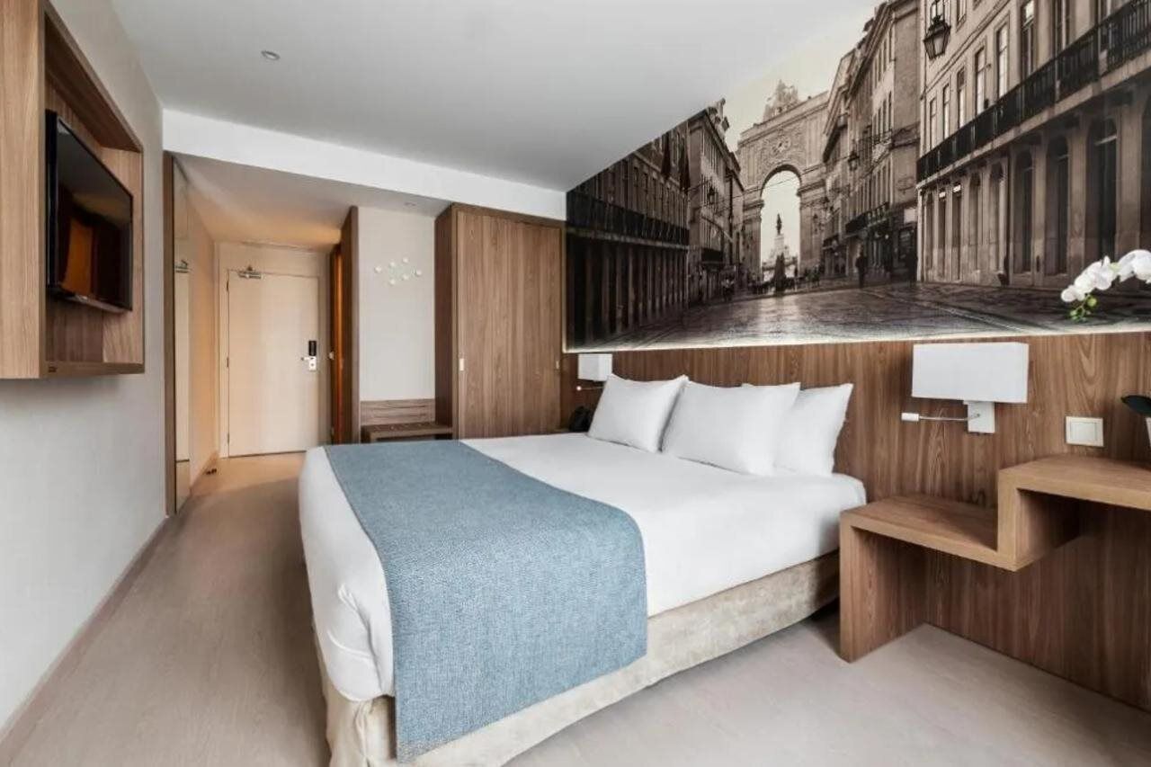 Quarto de hotel moderno com uma cama grande, cabeceira de madeira, televisão na parede e um mural de uma rua da cidade.