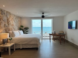 Quarto com cama, mesa de cabeceira, mesa e cadeira. Há uma janela ampla com vista para o mar.
