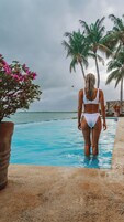 Mulher de biquíni branco em piscina, com palmeiras e vista para o oceano.
