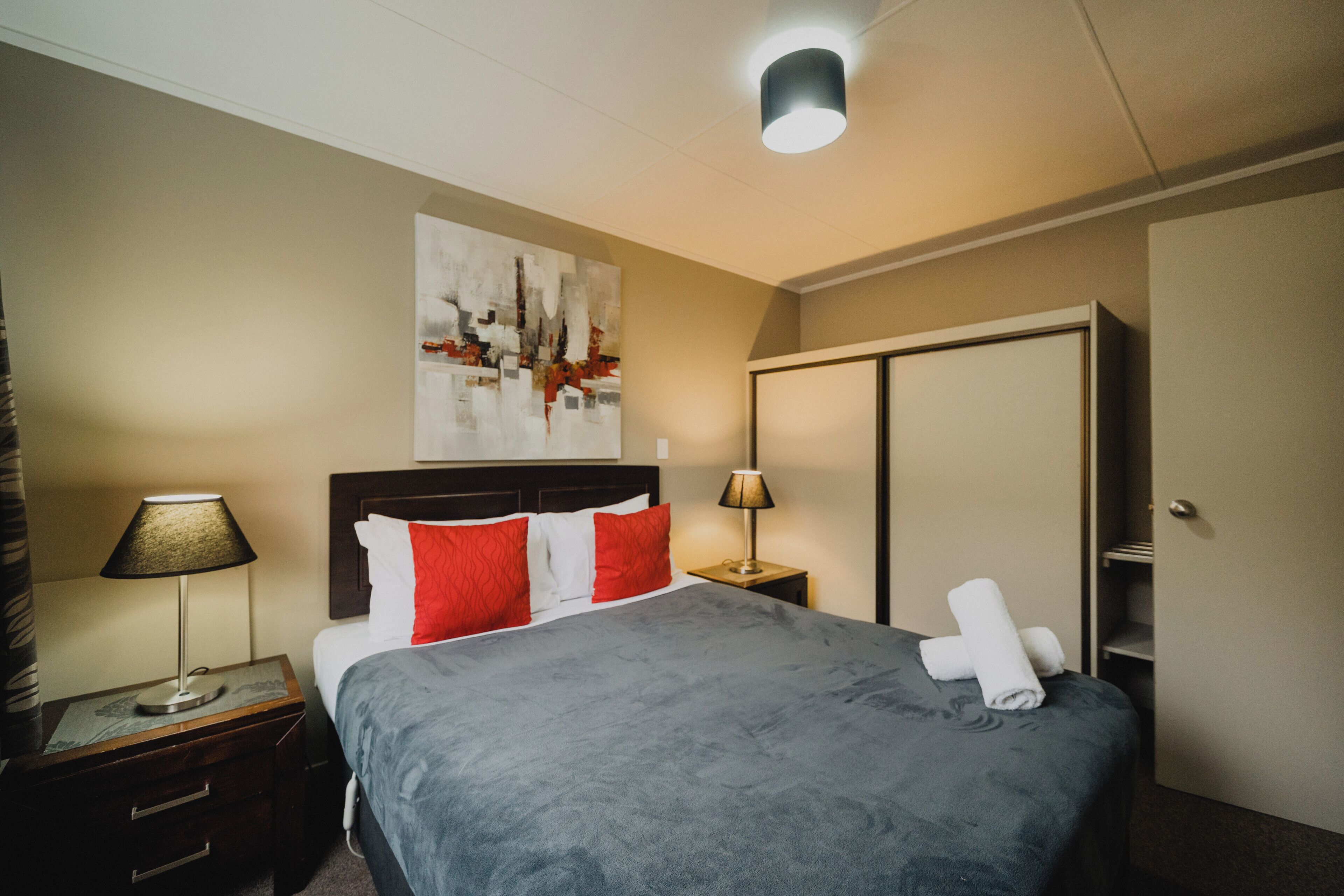 Quarto de hotel com cama, mesas de cabeceira, um quadro, guarda-roupa e uma luz no teto.