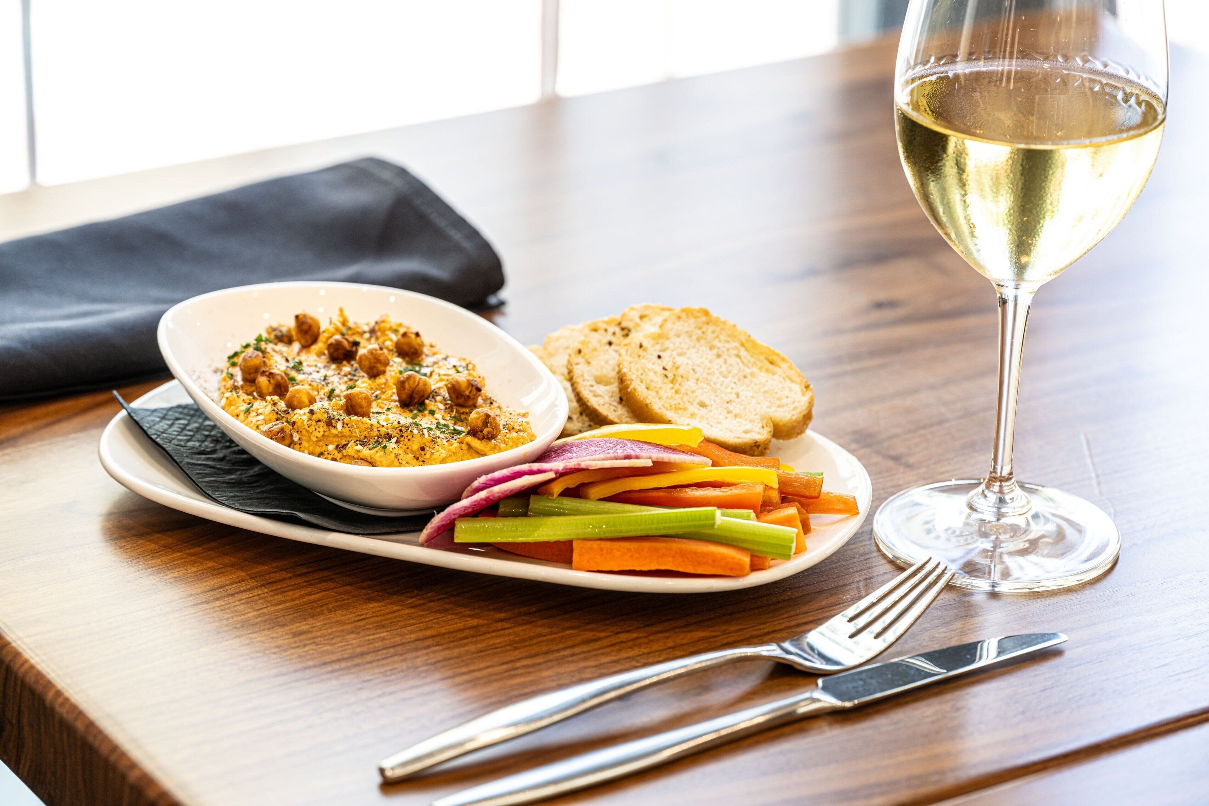 Uma refeição com uma tigela de sopa, legumes fatiados e pão, acompanhada por um copo de vinho branco sobre uma mesa de madeira.